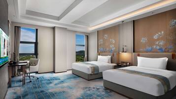 Quarto de hotel com duas camas, uma escrivaninha e uma cadeira. Janelas grandes oferecem vista para o exterior.