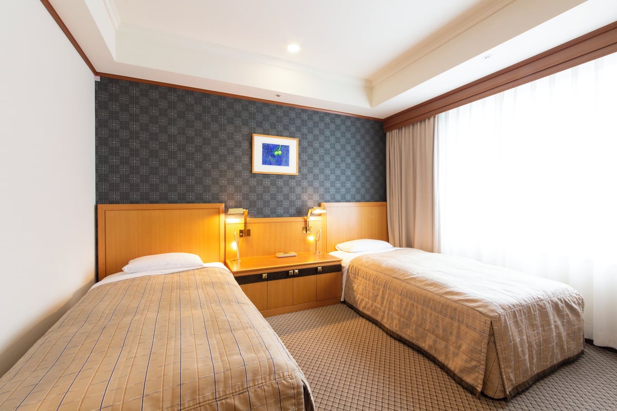 Quarto de hotel com duas camas, cabeceira, mesa de cabeceira, quadro emoldurado e janela com cortinas.
