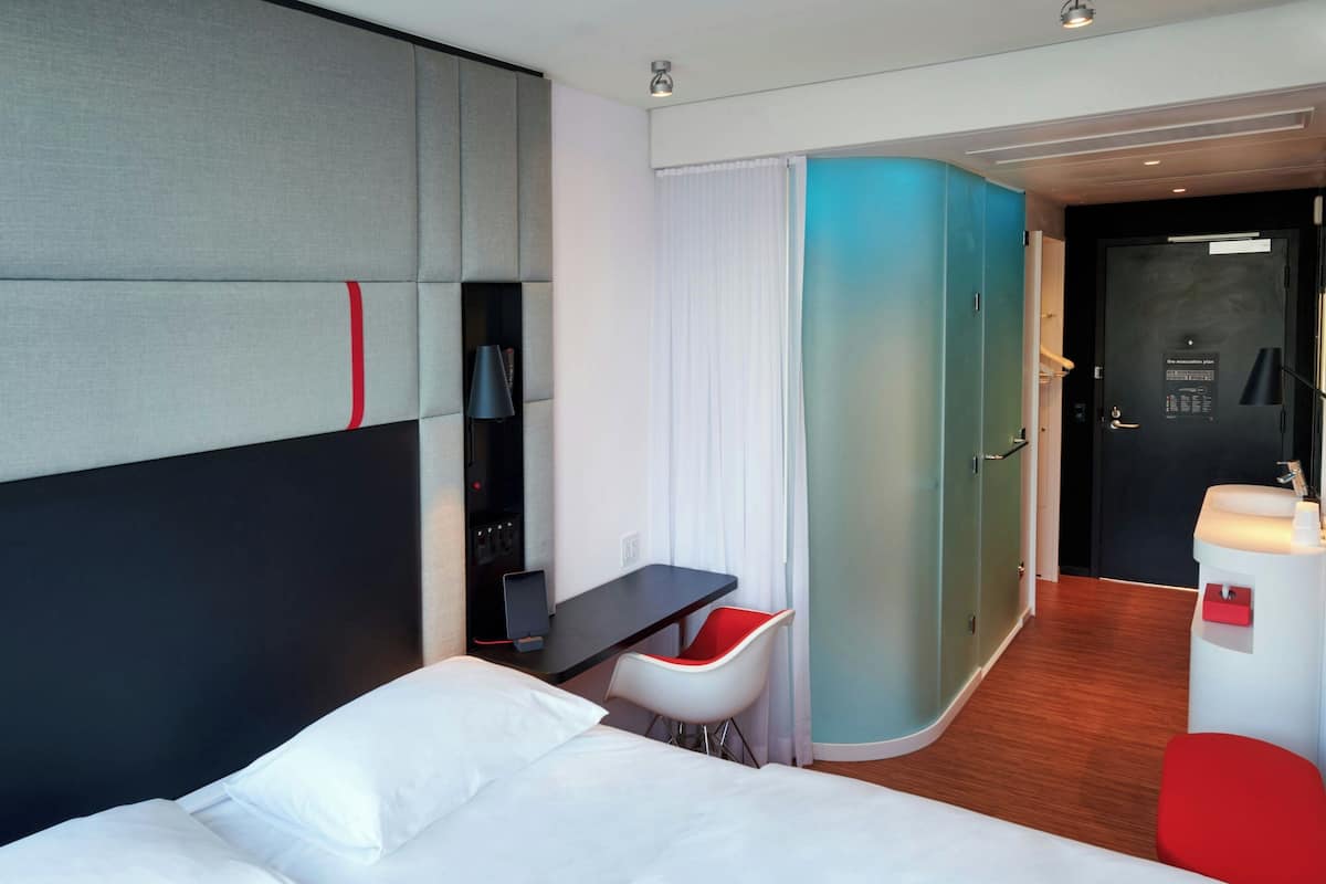 Quarto de hotel moderno com cama, escrivaninha, cadeira e banheiro.
