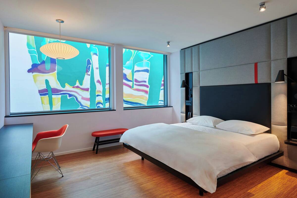 Quarto moderno com uma cama grande, um banco, uma escrivaninha e um quadro abstrato colorido nas janelas.