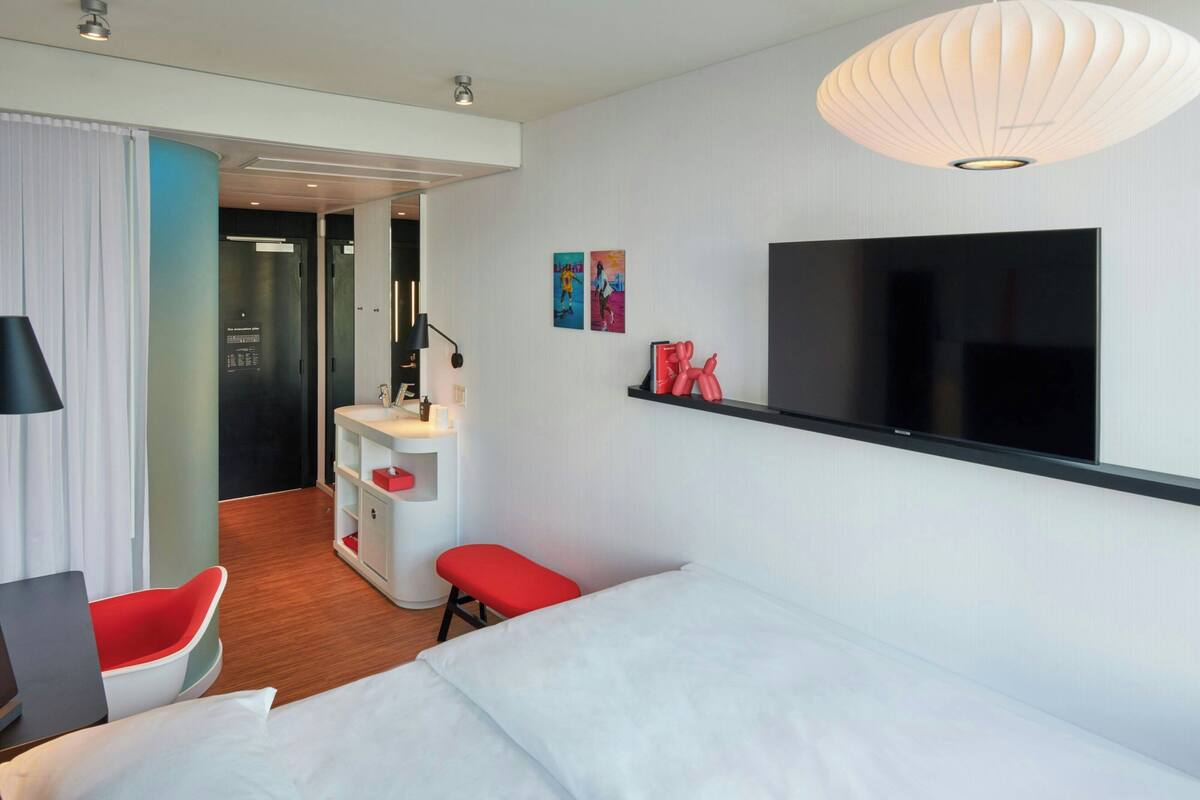 Quarto de hotel moderno com cama, escrivaninha com laptop, cadeira vermelha, toucador com espelho e televisão na parede.