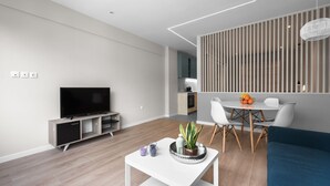 Uma sala de estar moderna com televisão de tela plana, uma mesa de centro branca com objetos decorativos, um sofá azul e um espaço de jantar com mesa e cadeiras brancas.