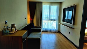 Quarto de hotel com piso de madeira, um sofá, uma mesa de centro com garrafas de água e uma cafeteira, uma TV fixada na parede e uma janela com cortinas translúcidas.
