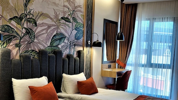 Quarto de hotel moderno com uma cama grande, uma escrivaninha e vista da cidade através de cortinas translúcidas.
