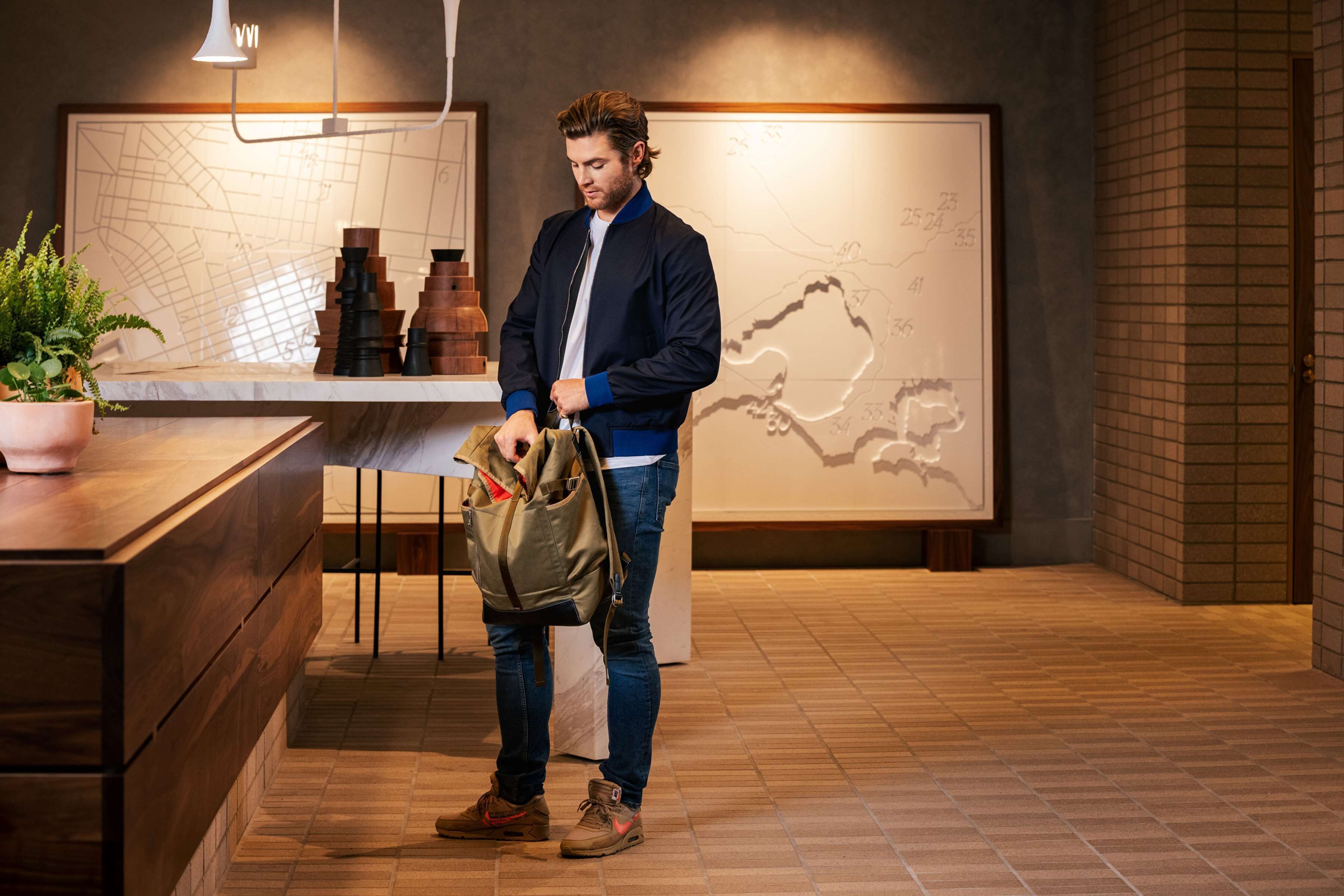 Um homem com casaco escuro e jeans está num saguão com um mapa na parede, um balcão de recepção de madeira e uma planta em vaso.