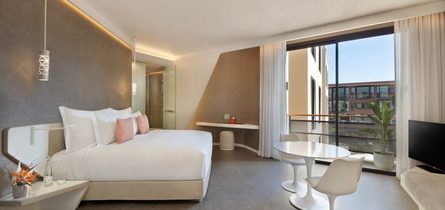 Quarto de hotel moderno com uma cama grande, uma varanda com televisão e um espaço de refeições.