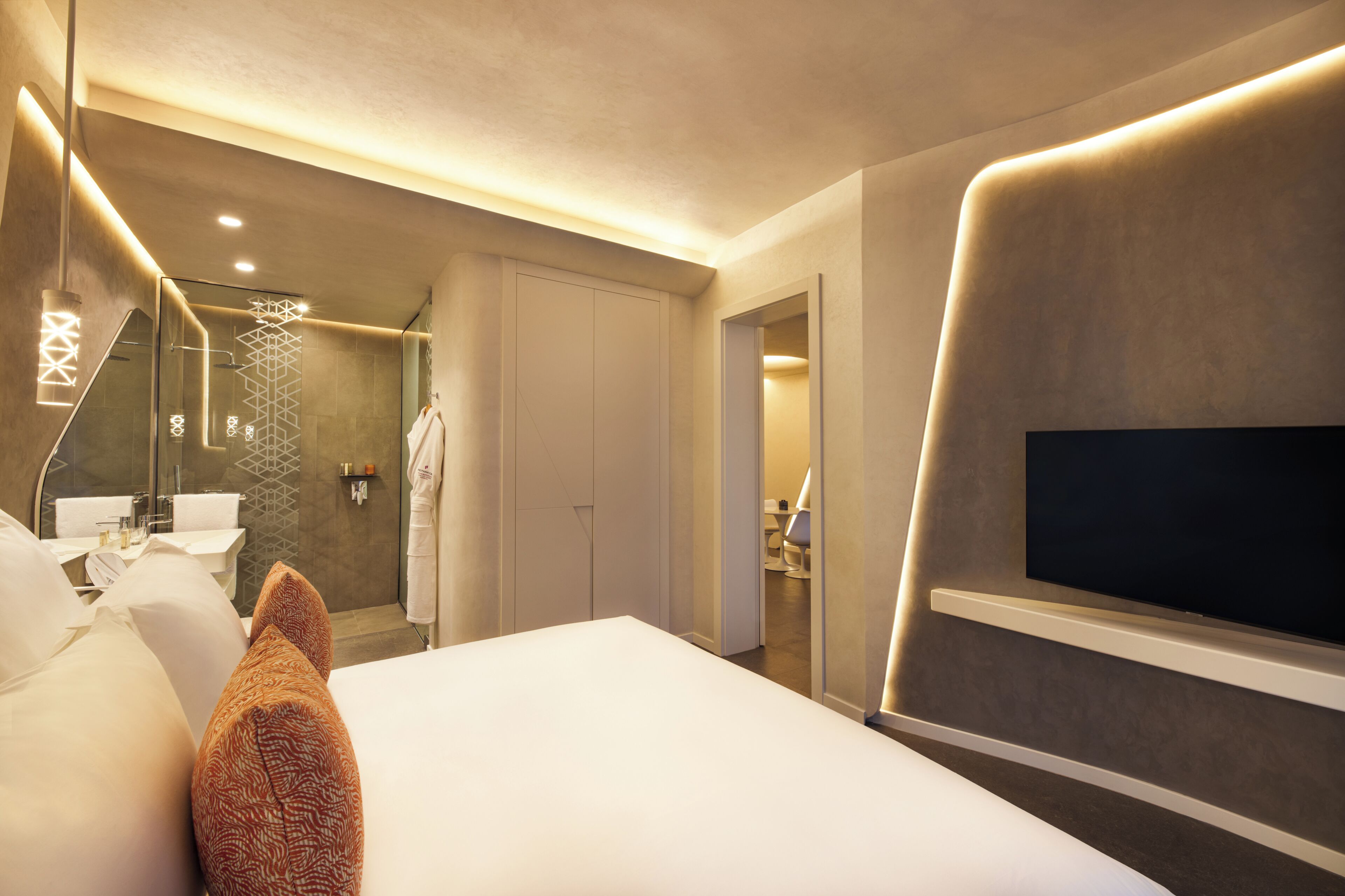 Quarto de hotel moderno com uma cama grande, uma TV fixada na parede, um banheiro com chuveiro de vidro e um closet espaçoso.