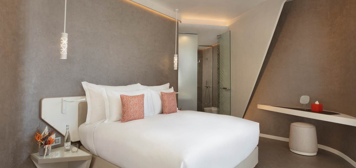 Quarto de hotel moderno com uma cama grande, mesas de cabeceira, um espelho e área de chuveiro.