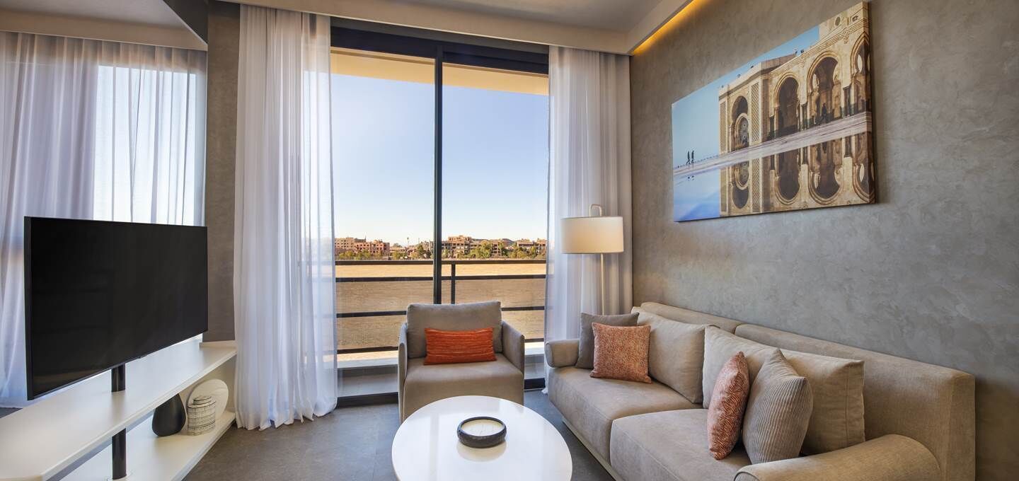 Quarto de hotel com uma televisão grande, sofá, mesa de centro e varanda com vista para a cidade.