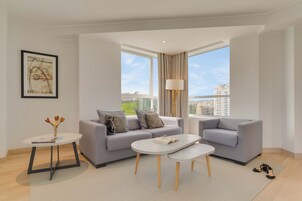 Uma sala de estar moderna com um sofá cinzento, duas mesinhas de centro brancas e vista para a cidade através de grandes janelas.