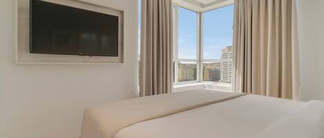 Quarto de hotel com cama, televisão fixada na parede e uma janela grande com cortinas.