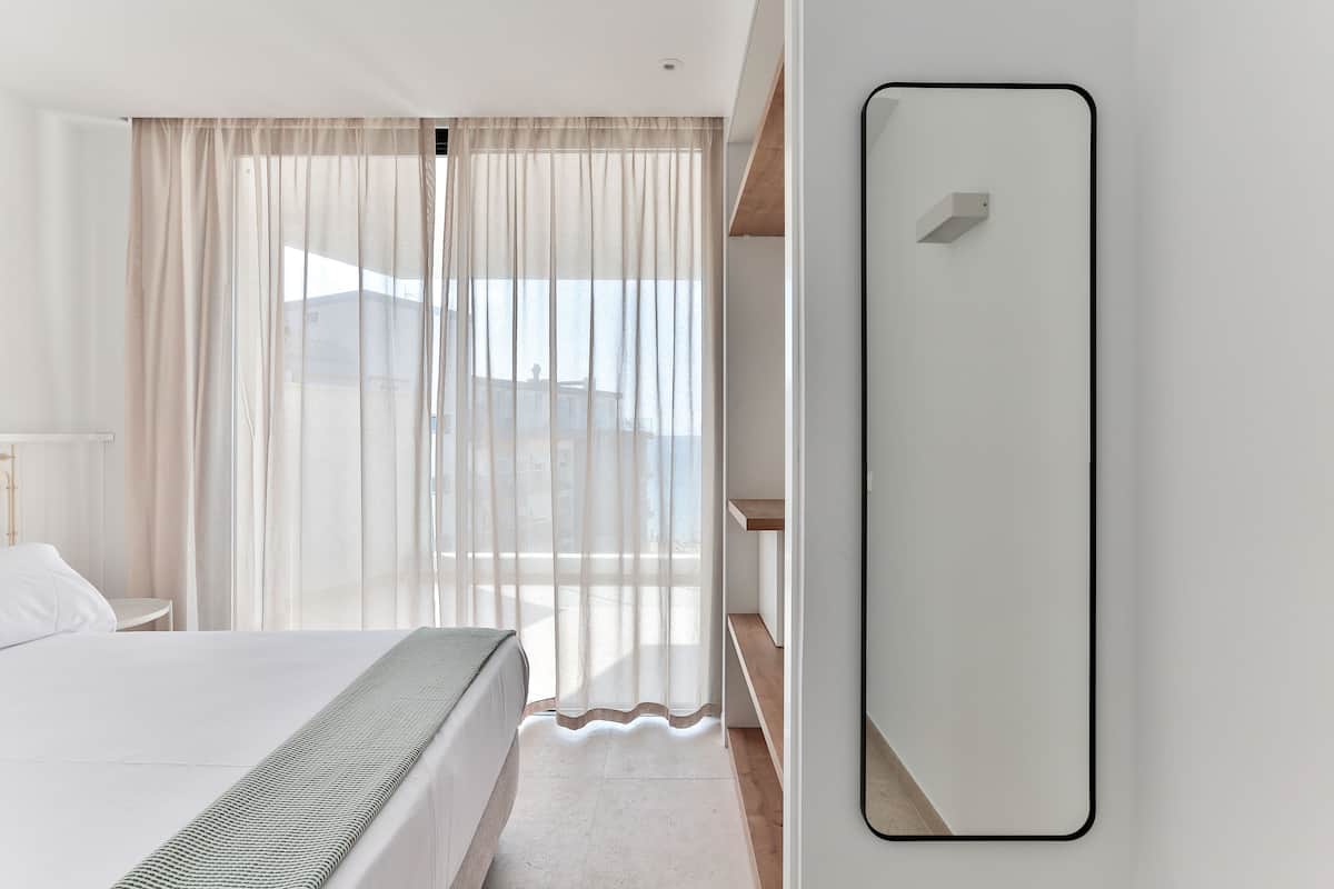 Quarto de hotel moderno com uma cama grande, uma mesa de cabeceira, um espelho e vista para o exterior através de cortinas translúcidas.