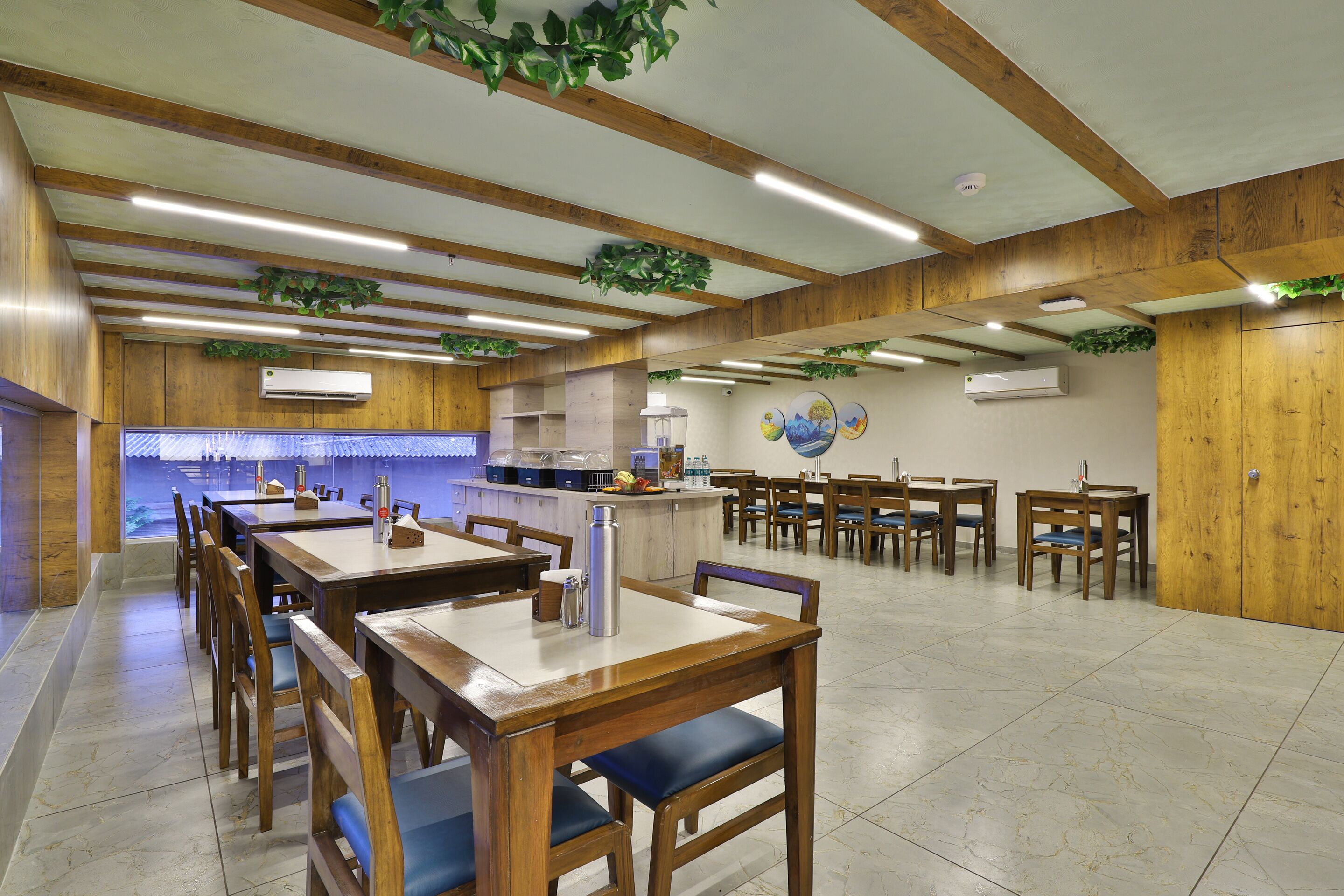 Uma sala de jantar ampla com mesas e cadeiras de madeira, um balcão central e plantas decorativas.