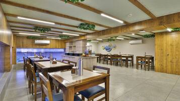 Uma sala de jantar ampla com mesas e cadeiras de madeira, um balcão central e plantas decorativas.