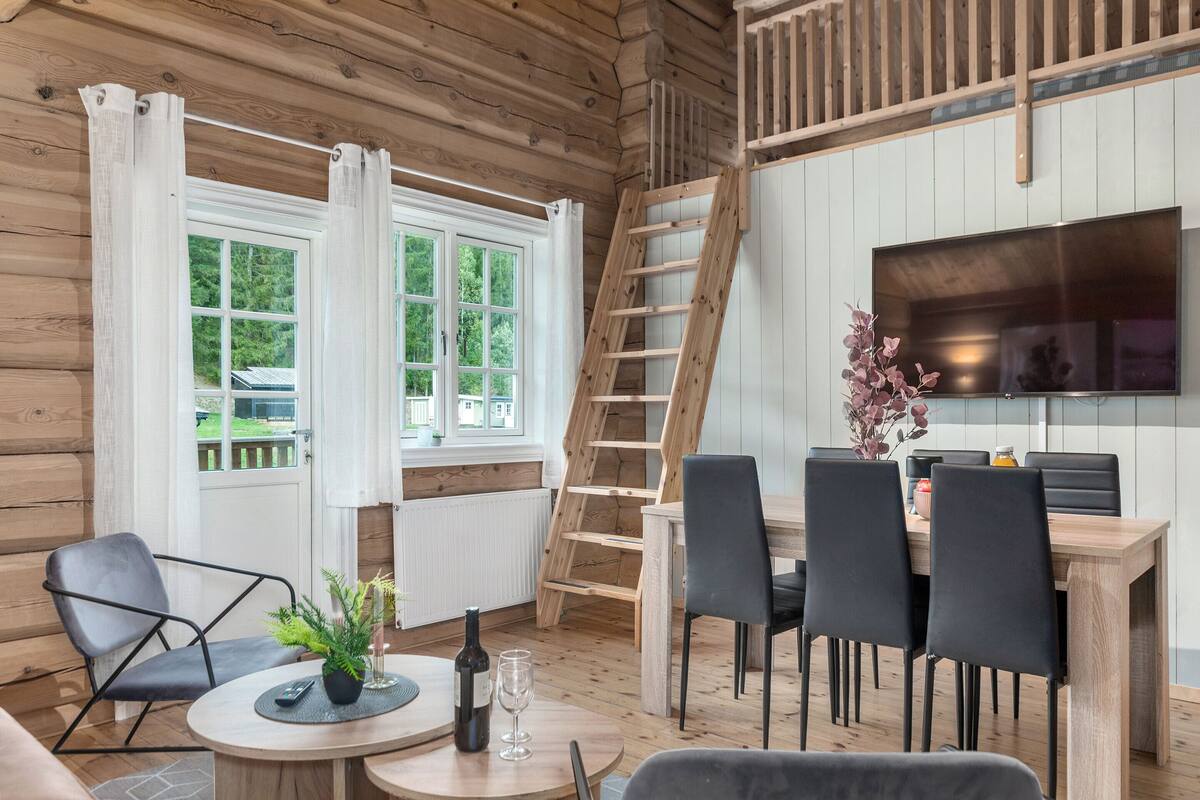 Interior acolhedor de uma cabana de madeira, com área de jantar, TV fixada na parede e escada de madeira para um mezanino.