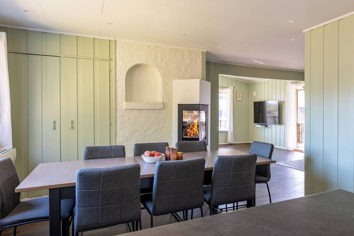 Uma área de jantar moderna com uma mesa central, cadeiras estofadas cinzentas, lareira e televisão na parede.