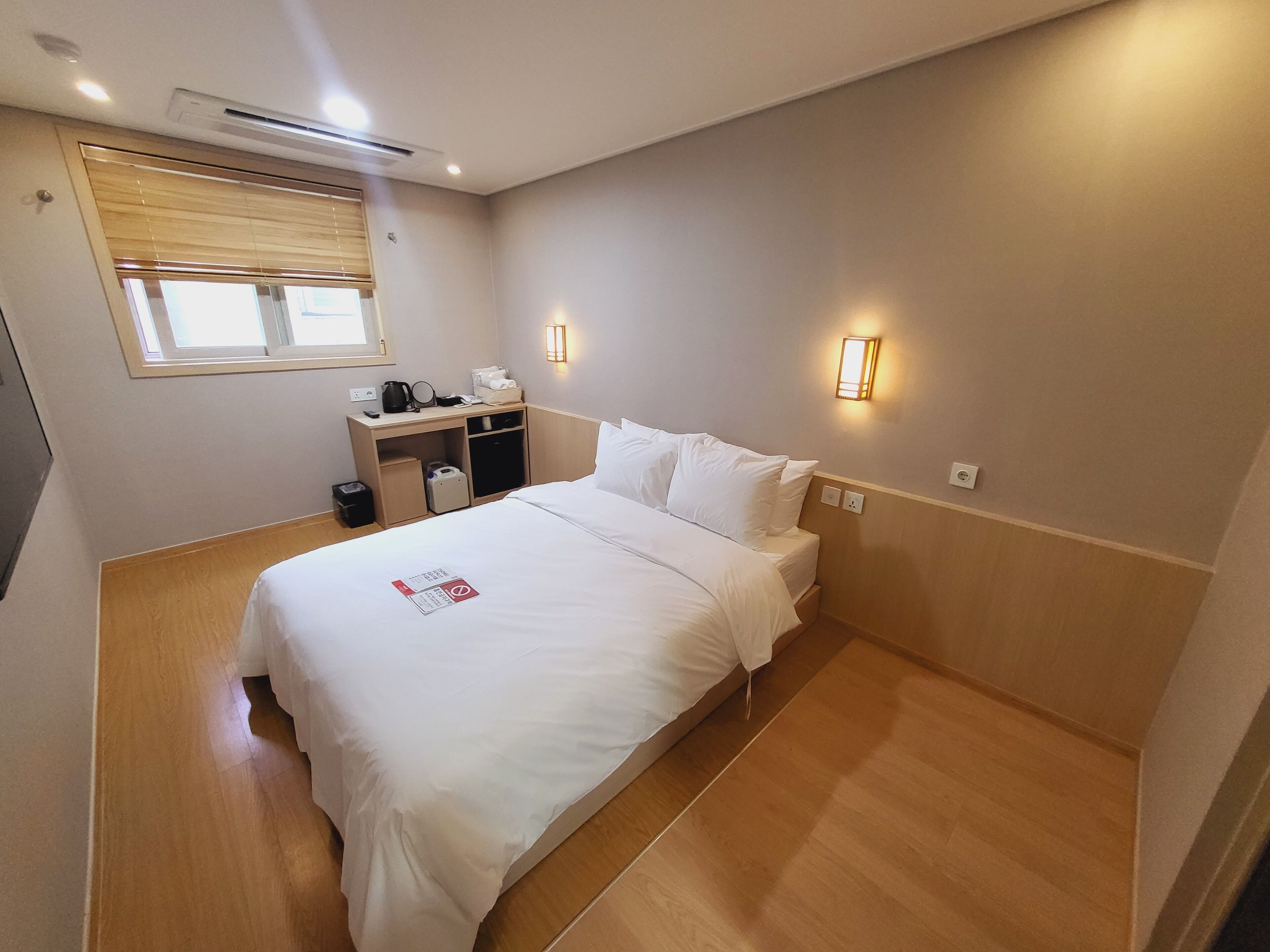 Quarto de hotel com cama grande, uma escrivaninha com cadeira, guarda-roupa pequeno e janela com persianas.