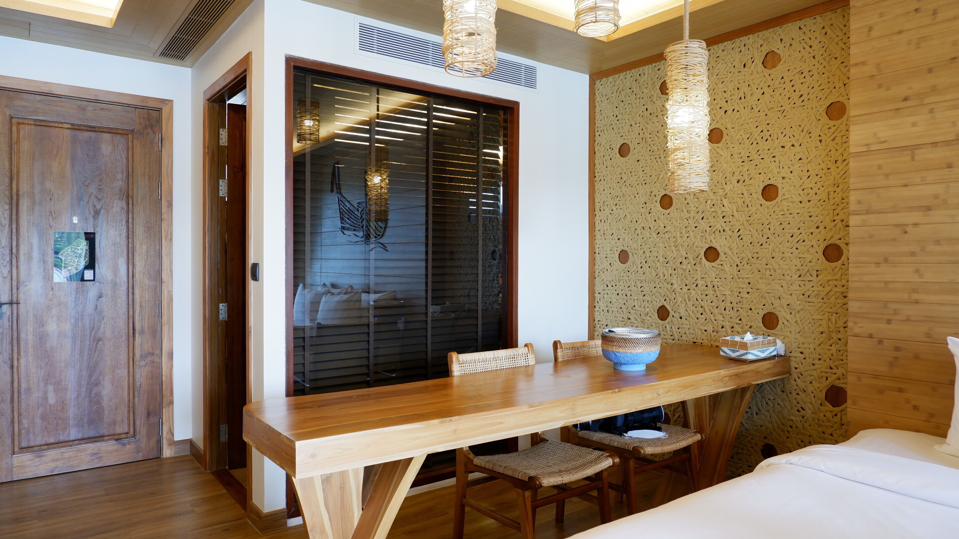 Uma mesa de jantar de madeira com cadeiras, uma janela grande com persianas e um painel de parede trançado.