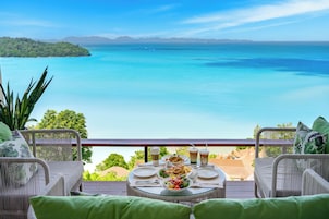 Mesa de jantar com pratos, talheres e bebidas em uma varanda com vista para um mar azul-turquesa e paisagem verde.