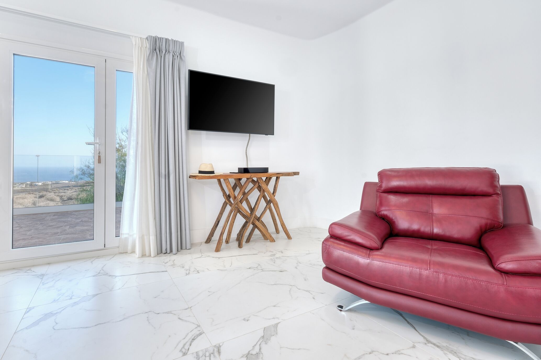 Uma sala de estar moderna com um sofá de couro vermelho, uma TV de tela plana na parede, uma mesa de centro de madeira e uma grande porta de vidro deslizante que dá para uma varanda com vista para o mar.