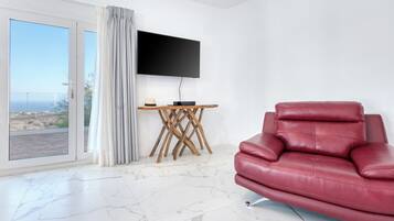 Uma sala de estar moderna com um sofá de couro vermelho, uma TV de tela plana na parede, uma mesa de centro de madeira e uma grande porta de vidro deslizante que dá para uma varanda com vista para o mar.