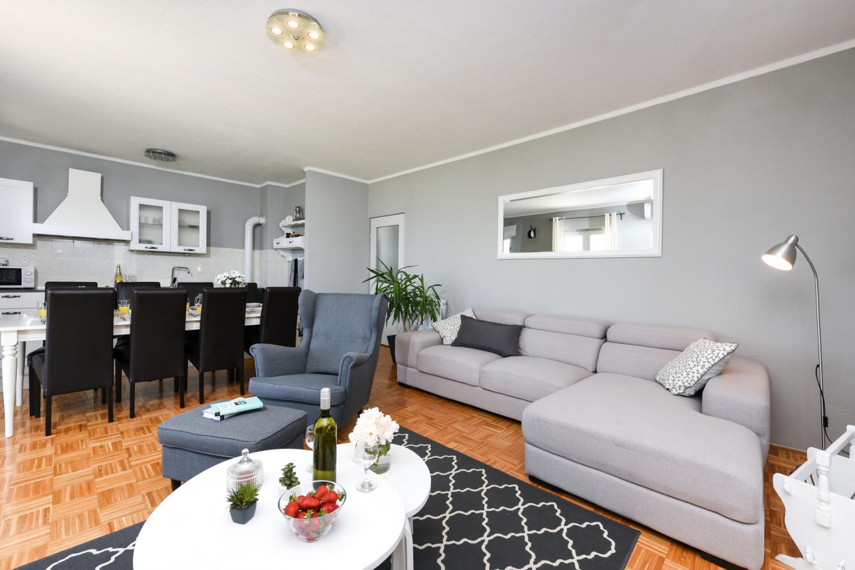Uma sala de estar moderna com sofá cinzento, área de jantar com cadeiras pretas, mesa de centro branca com flores e uma garrafa, e uma cozinha ao fundo.
