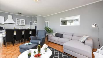 Uma sala de estar moderna com sofá cinzento, área de jantar com cadeiras pretas, mesa de centro branca com flores e uma garrafa, e uma cozinha ao fundo.
