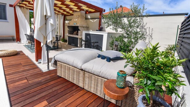 Um deck de madeira com sofá de vime, área de estar externa, pérgula e espaço para churrasco.
