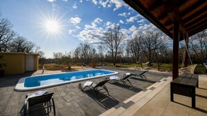 Piscina com espreguiçadeiras e mesa sob uma área coberta, cercada por árvores e um céu claro.