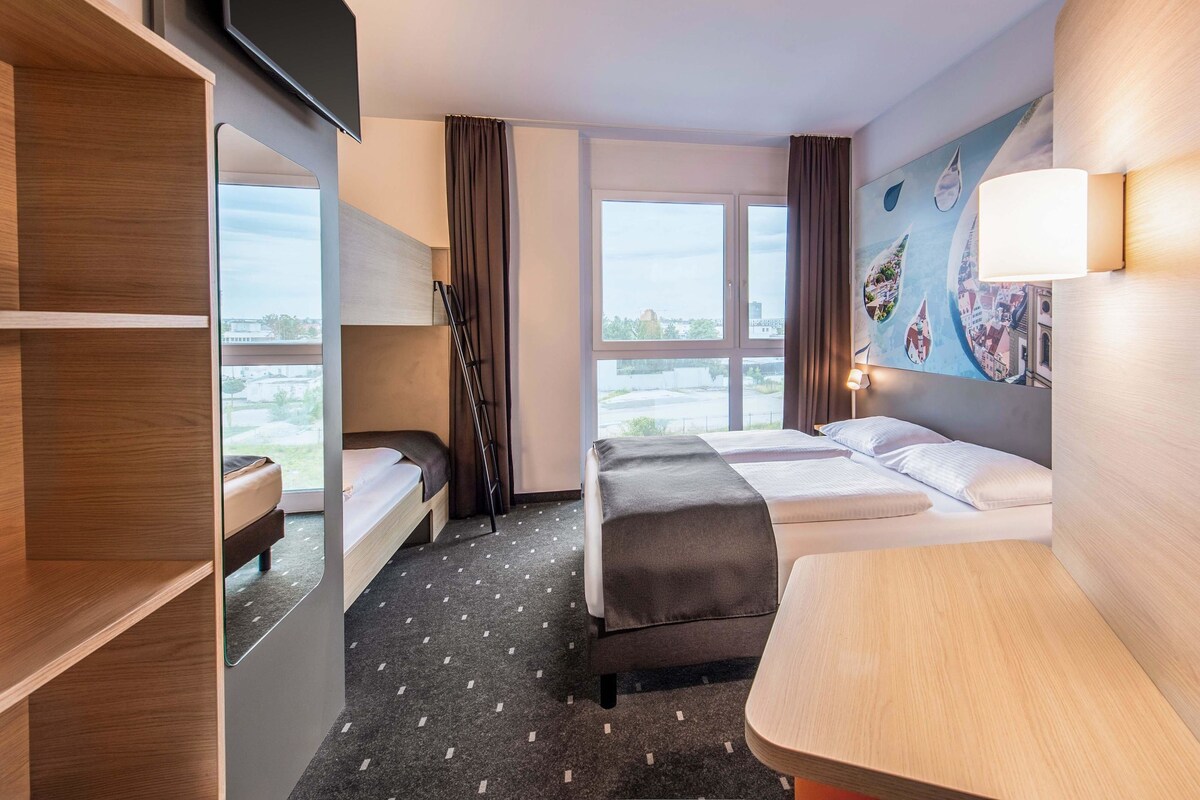 Quarto de hotel com cama beliche, uma escrivaninha, uma cadeira, televisão e janela com vista para a cidade.