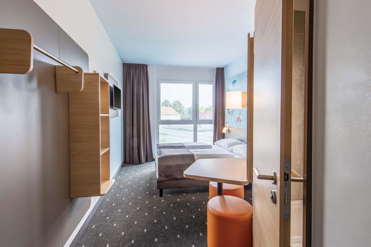 Quarto de hotel com cama, escrivaninha, cadeira, televisão e janela com vista para o exterior.