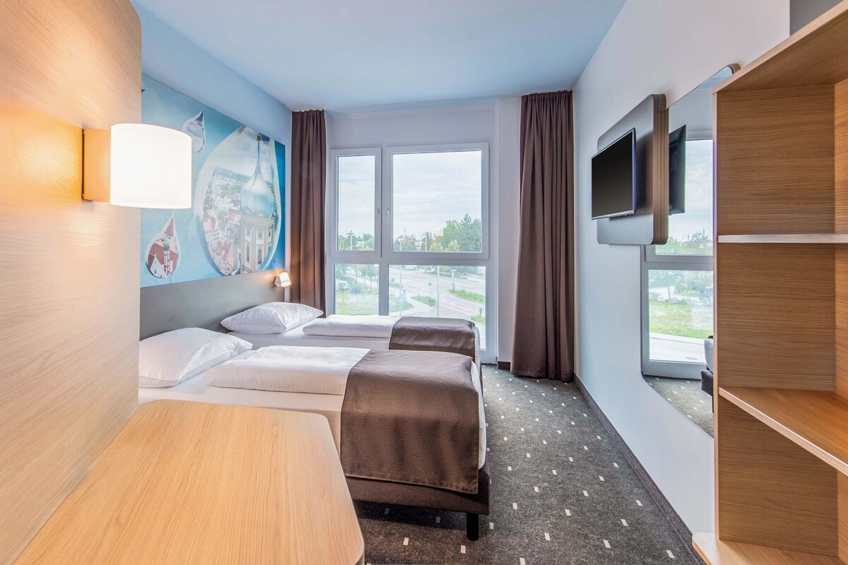 Quarto de hotel com duas camas, uma escrivaninha, televisão e uma janela grande com vista para a cidade.