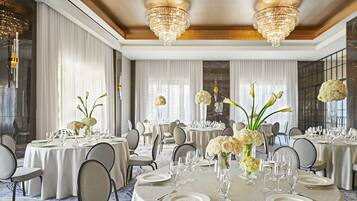 Uma sala de jantar formal com mesas redondas preparadas para o serviço, elegantes lustres e amplas janelas com cortinas.
