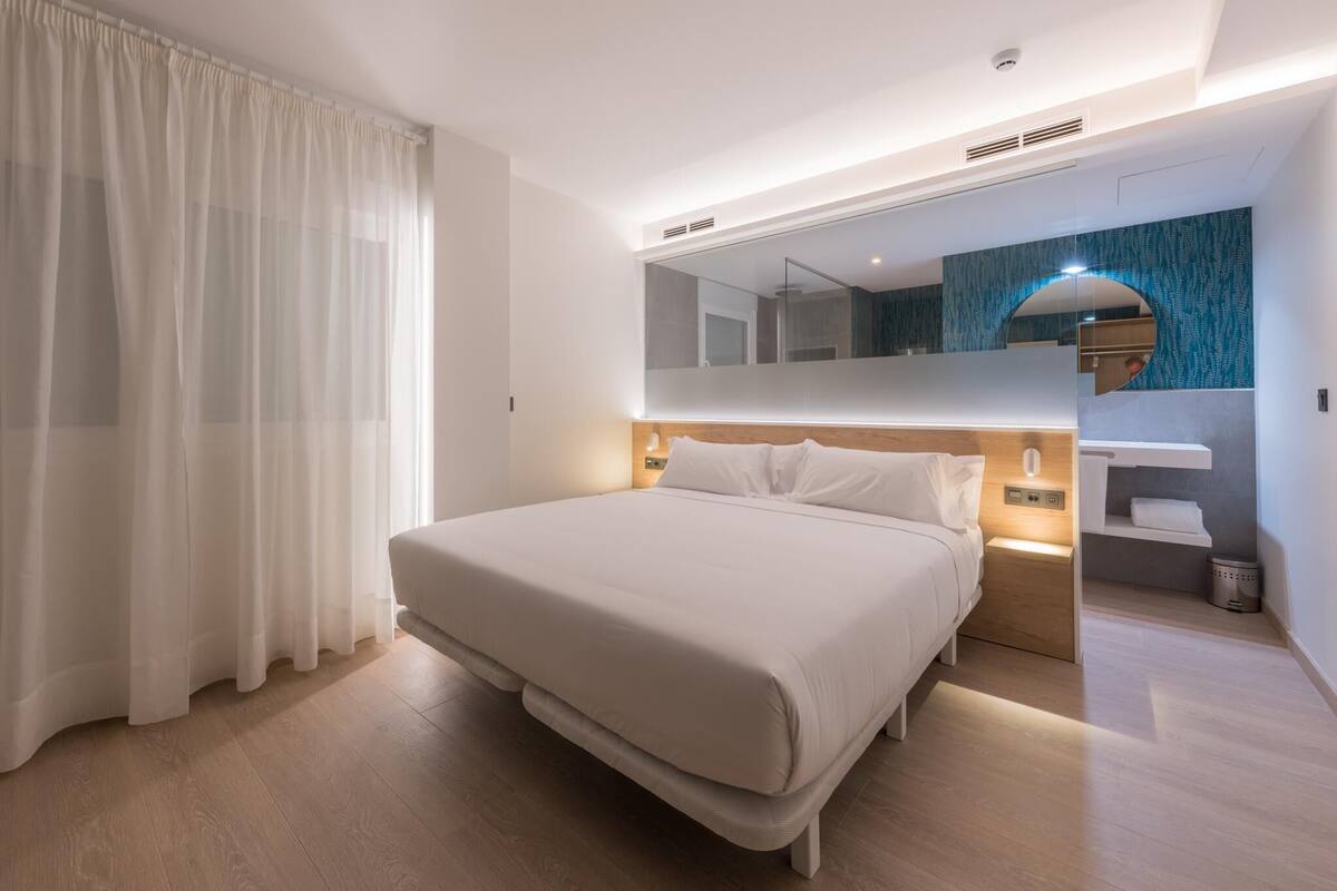 Quarto de hotel moderno com uma cama grande, uma mesinha de cabeceira, um espelho e uma pia pequena.