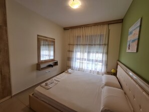 Quarto com cama grande, mesa de cabeceira, espelho, janela com cortinas e um quadro na parede.