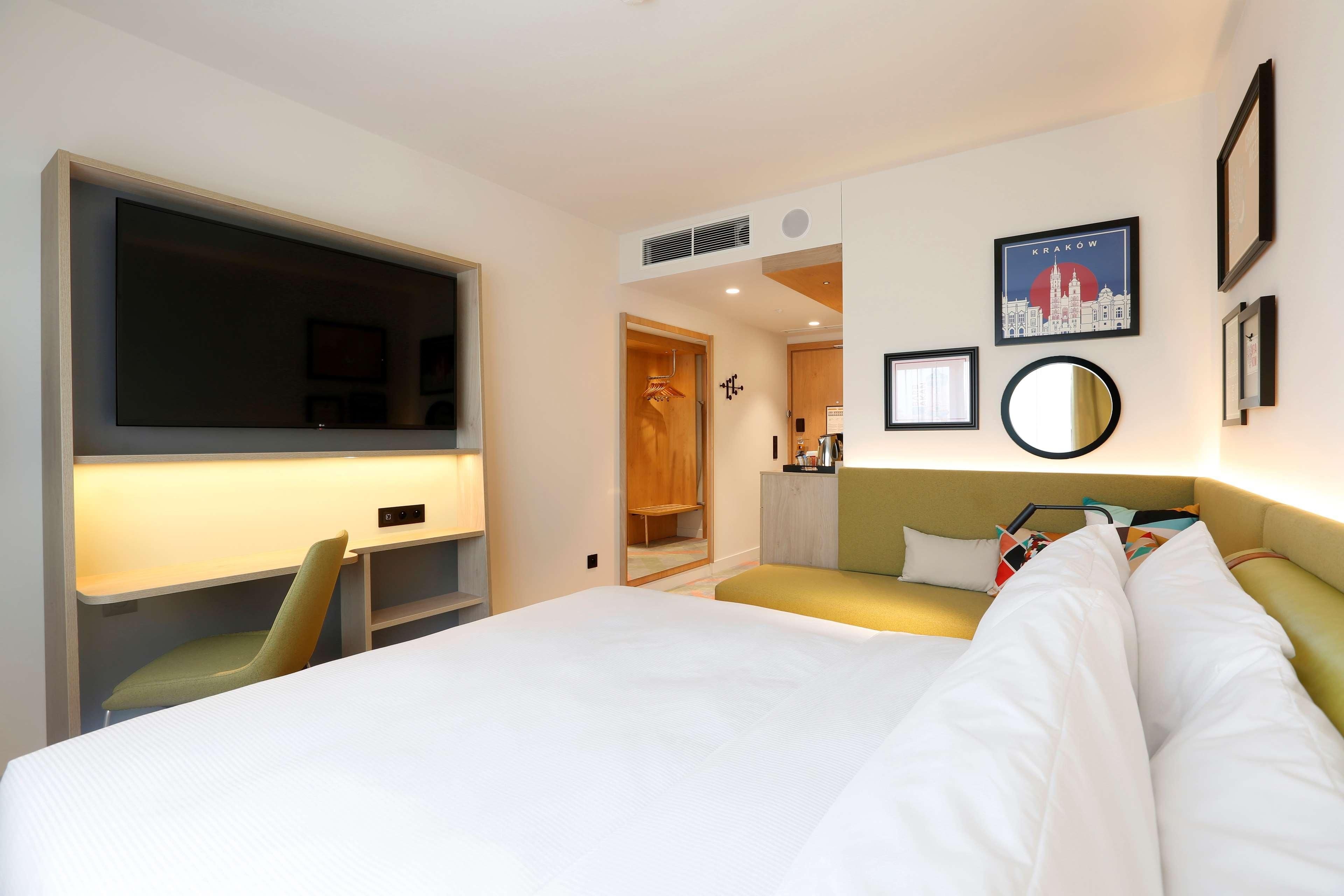 Quarto de hotel com cama, escrivaninha, cadeira, televisão, guarda-roupa e espelho.