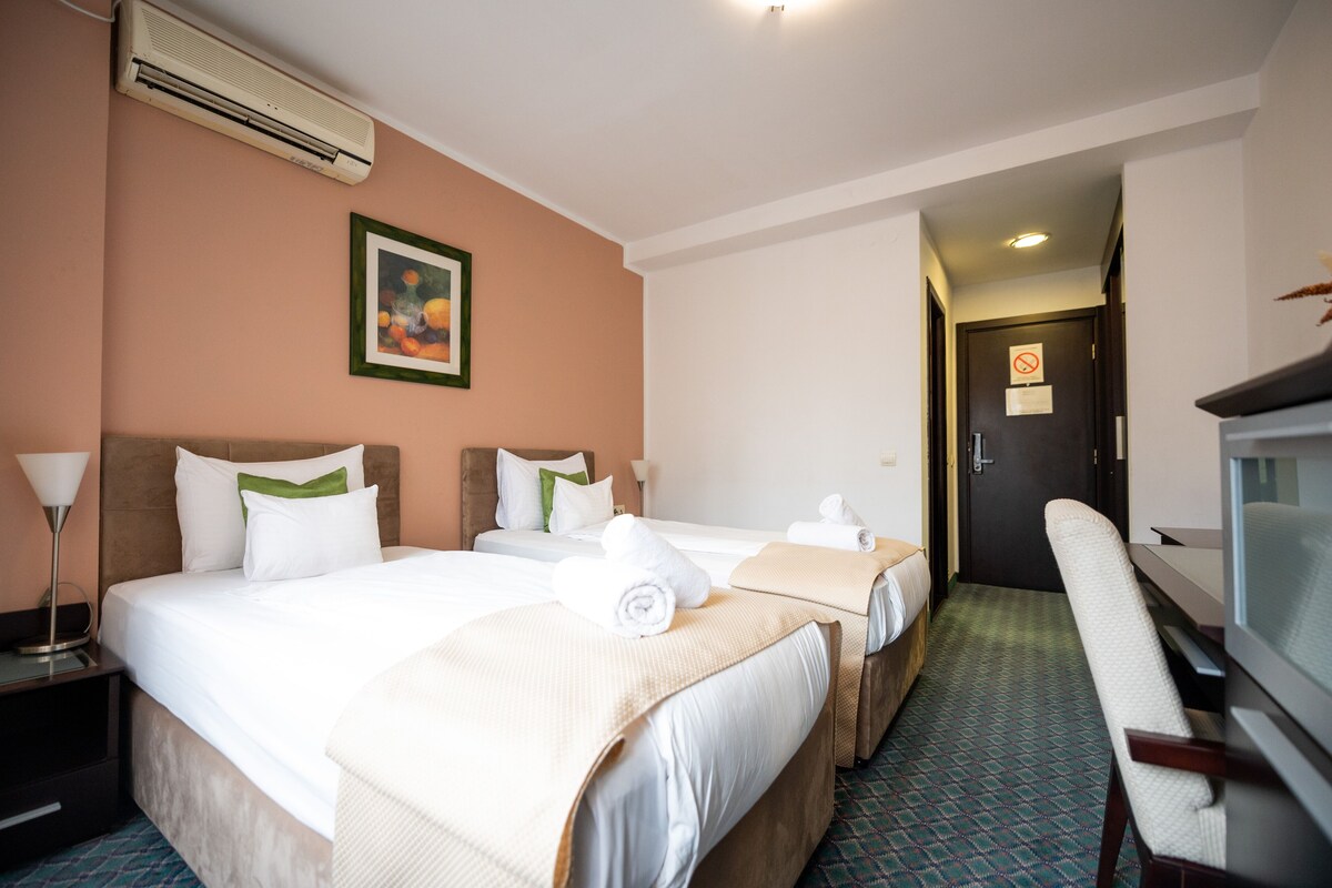 Quarto de hotel com duas camas, televisão, escrivaninha e porta com aviso de proibido fumar.
