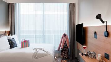 Quarto de hotel com cama, televisão, escrivaninha com luminária, janela com cortinas e vista para a cidade.