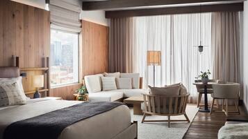 Quarto de hotel moderno com uma cama grande, um sofá, uma cadeira, uma mesa pequena e vista para a cidade.