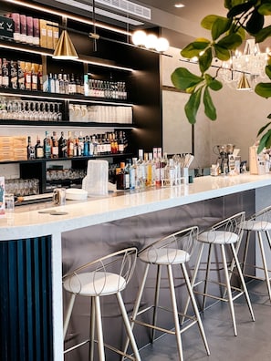 Uma área de bar com um balcão comprido, várias prateleiras com garrafas de bebidas e três banquetas de metal.