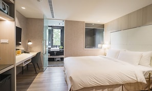 Quarto de hotel moderno com uma cama grande, uma escrivaninha e um banheiro visível através de uma porta de vidro.