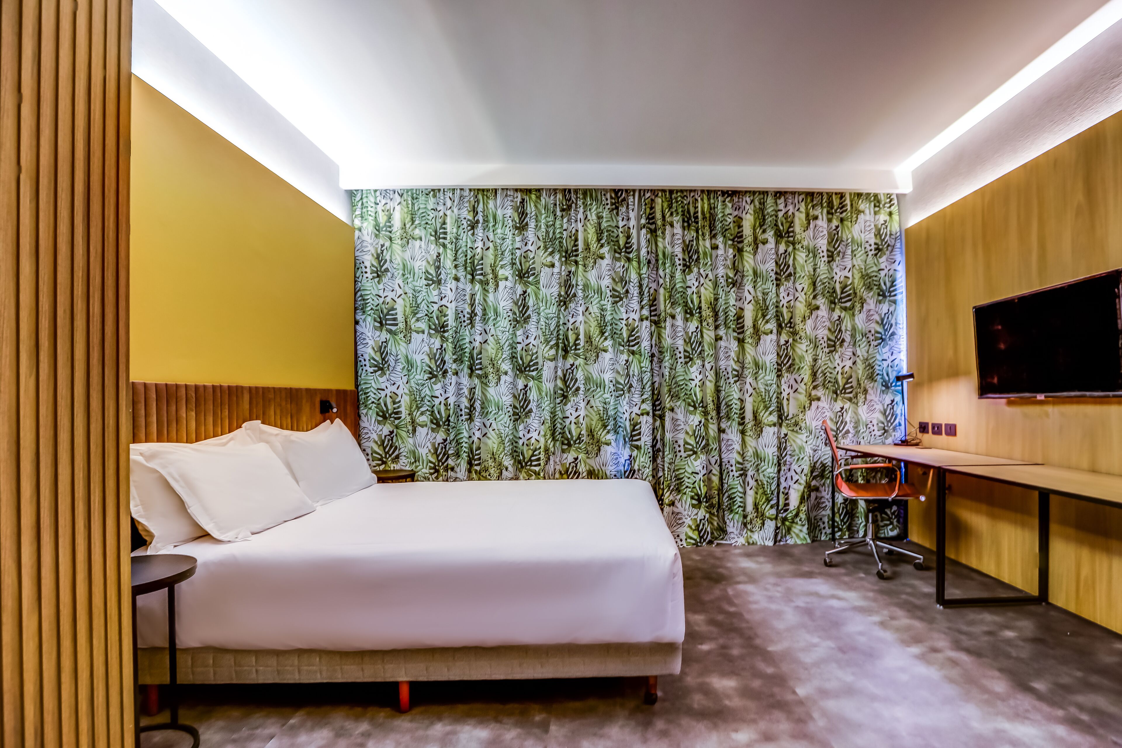 Quarto de hotel moderno com cama, escrivaninha, cadeira e televisão.