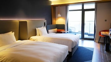 Quarto de hotel com duas camas, uma mesinha, uma cadeira e vista para o exterior.