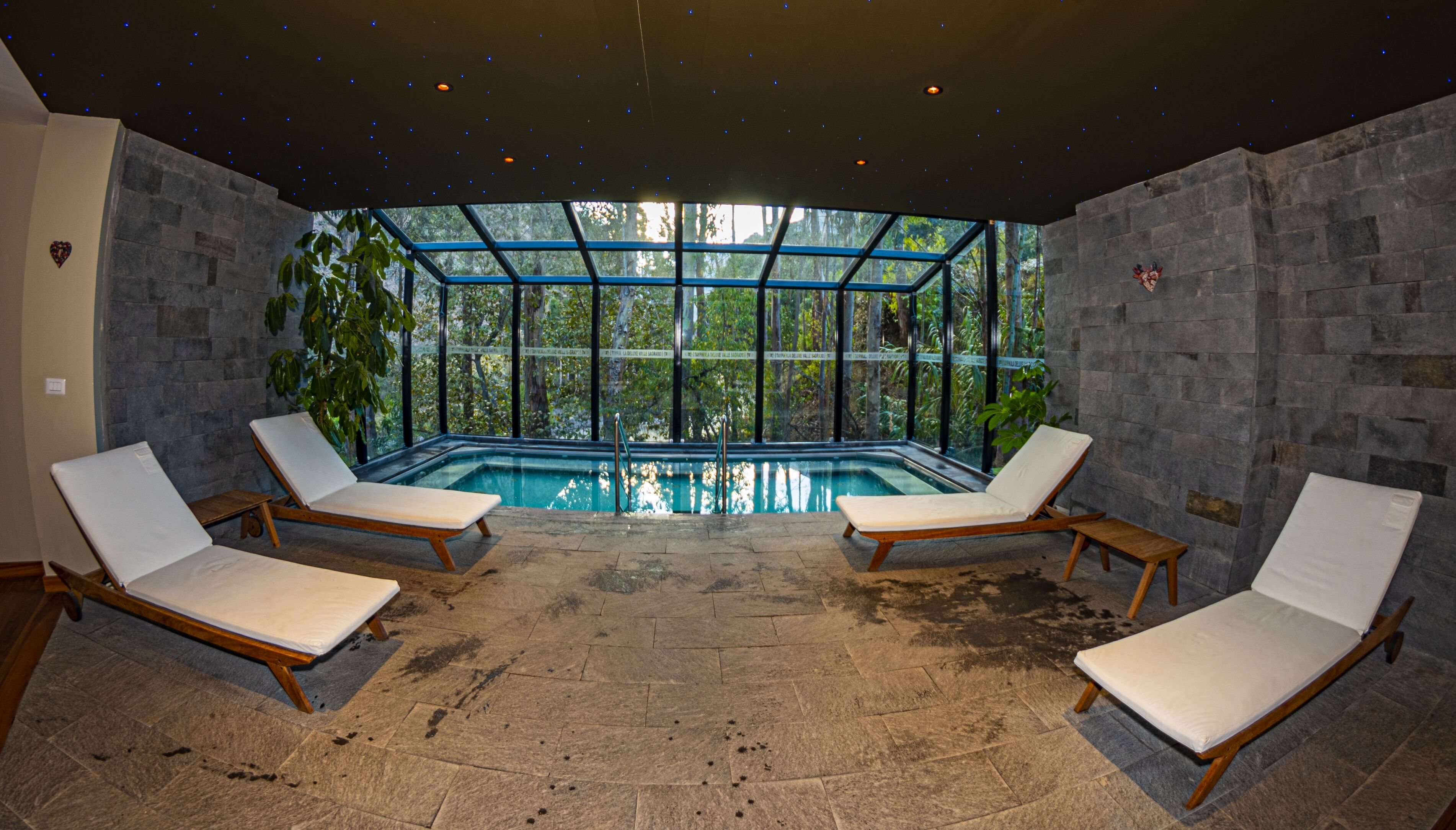 Uma área de spa com piscina, duas espreguiçadeiras, paredes de pedra e vista para árvores através de grandes janelas.