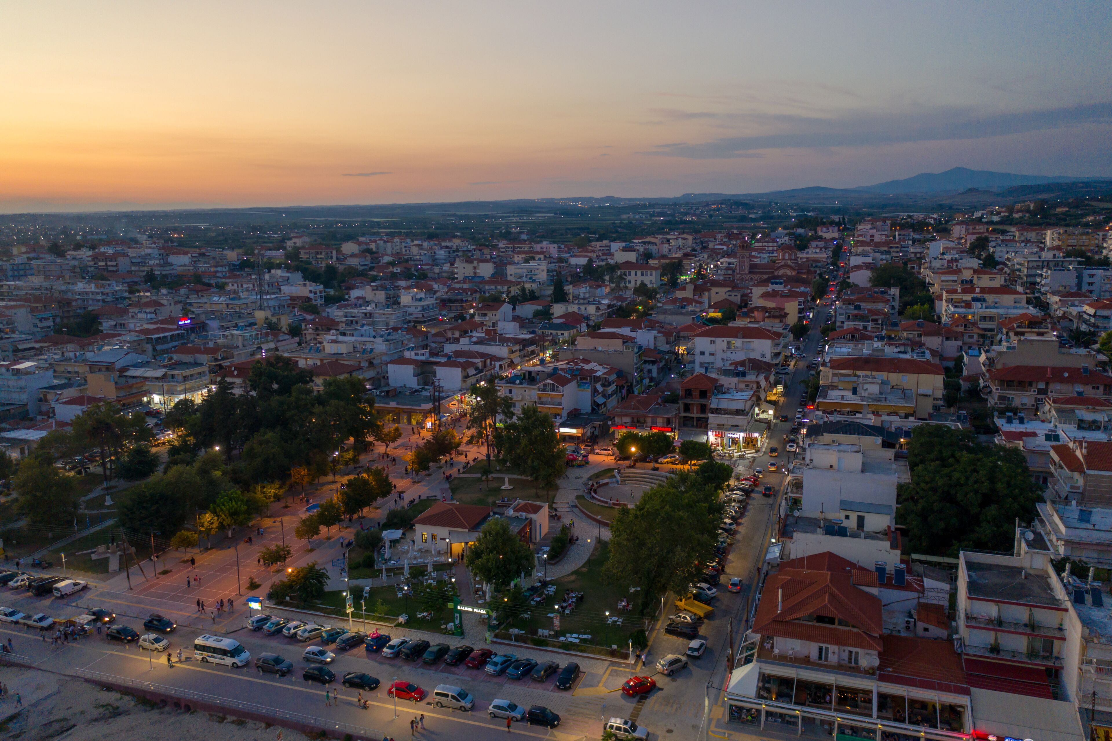 Vista aérea de um horizonte urbano ao entardecer, com mistura de edifícios residenciais e comerciais, um cruzamento rotatório central e uma área de estacionamento.
