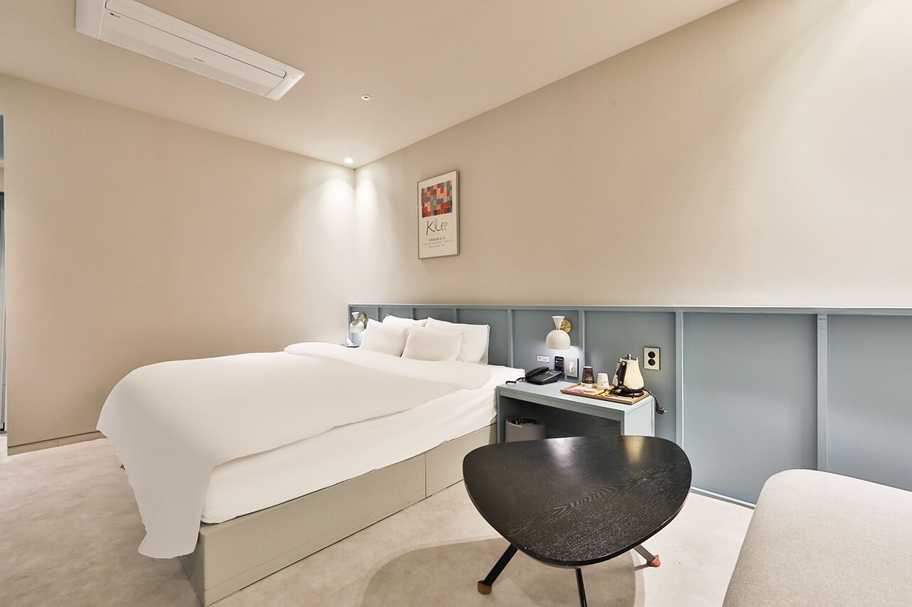 Quarto de hotel com cama, escrivaninha com luminária, cadeira e pequena mesa com telefone e luminária.