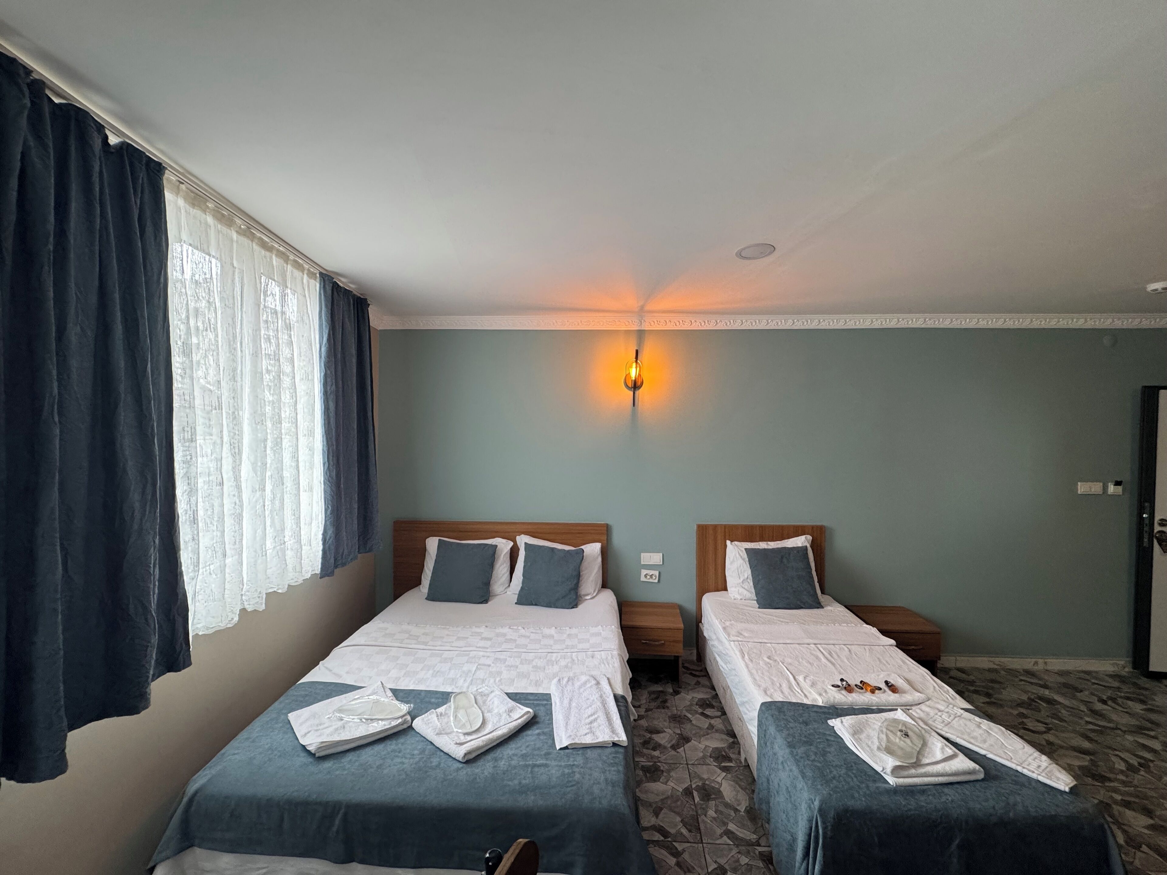 Quarto de hotel com duas camas, mesa de cabeceira, abajur e janela com cortinas.