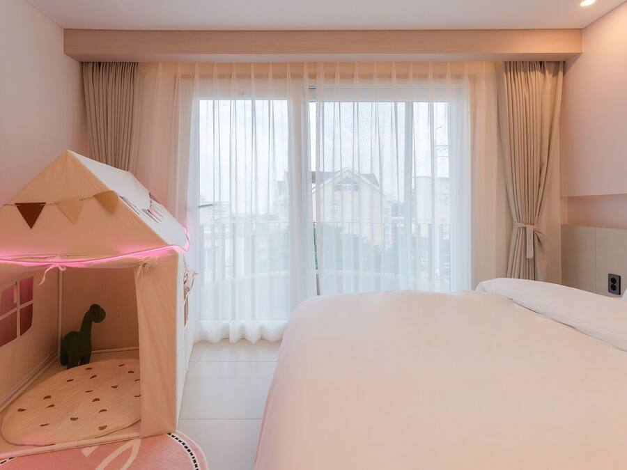 Quarto de hotel com cama, uma tenda de brincar, TV fixada na parede e janela com cortinas.