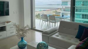Uma sala de estar moderna com sofá, mesa de centro e televisão. Uma janela grande oferece vista para uma praia e um edifício do outro lado da água.