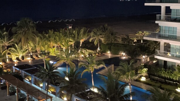 Um hotel moderno com área de piscina, palmeiras e vista para a praia à noite.